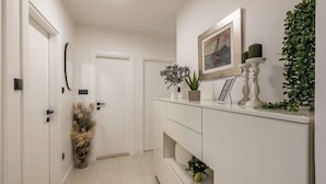 Corredor com paredes brancas, porta branca, um console com uma moldura e uma planta em vaso, além de um vaso com flores.