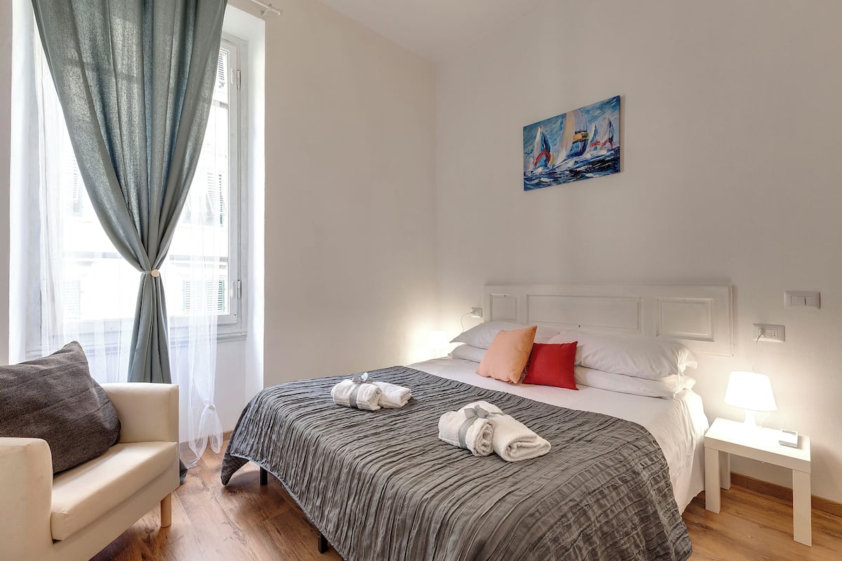 Quarto de hotel com cama, mesa de cabeceira com abajur, uma cadeira e um quadro na parede.