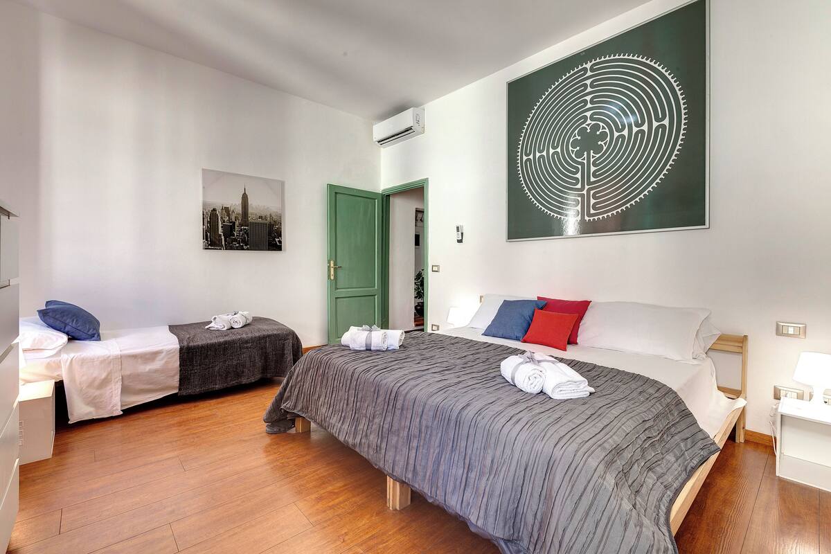 Quarto de hotel com uma cama grande, uma cama menor, uma porta verde e um ar condicionado fixado na parede.