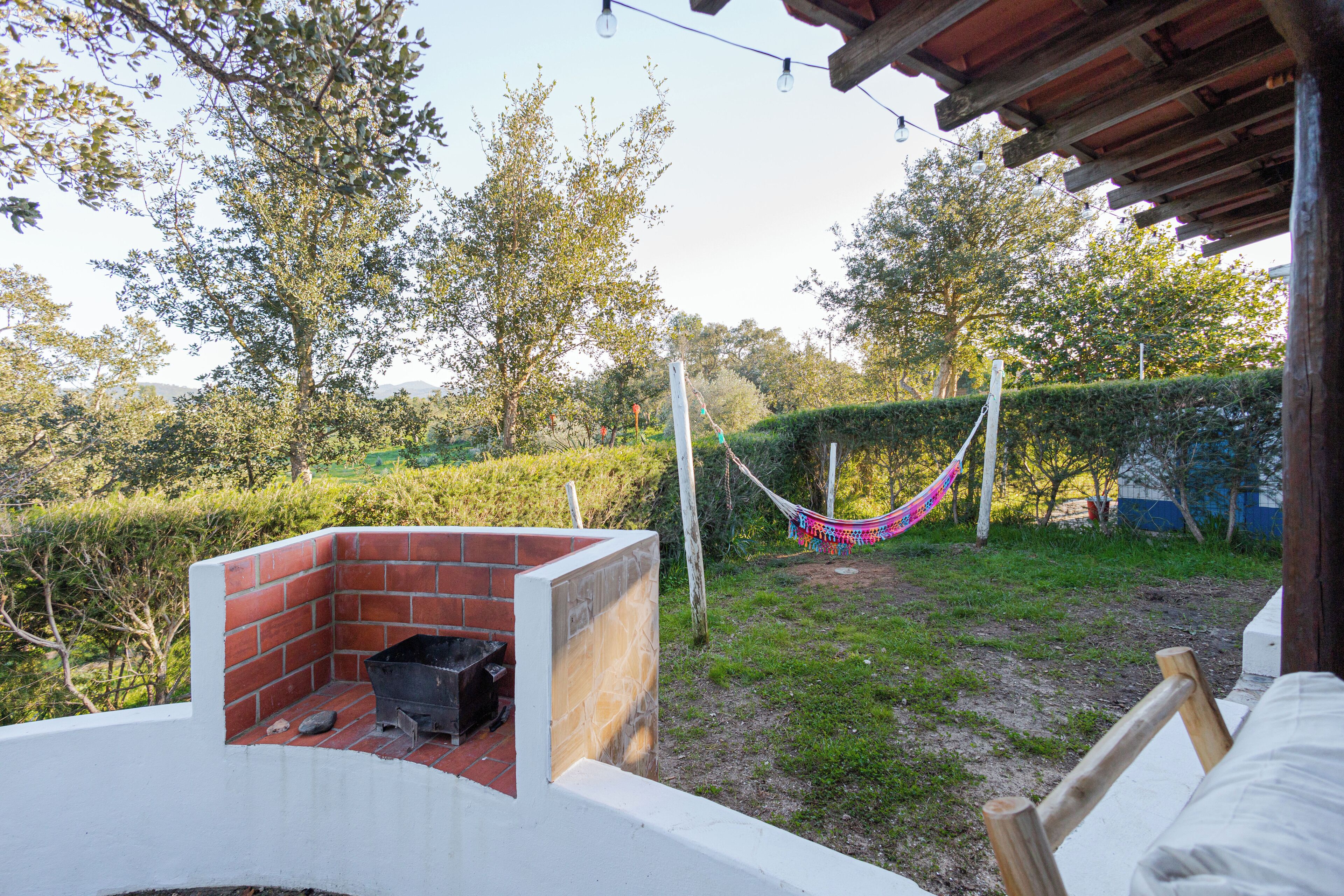 Uma área de pátio com churrasqueira de tijolos, uma rede e vista para o jardim e árvores ao redor.
