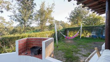 Uma área de pátio com churrasqueira de tijolos, uma rede e vista para o jardim e árvores ao redor.
