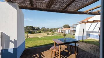 Um deck de madeira com uma mesa e um banco, com vista para uma área gramada e edifícios ao fundo.