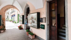 Uma entrada em arco com uma placa indicando “I PORTICI”, uma vitrine de vidro com objetos, uma caixa de correio e plantas em vasos.