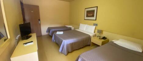 Quarto de hotel com duas camas de solteiro, uma escrivaninha com computador, um telefone e uma pequena mesa de cabeceira com abajur.