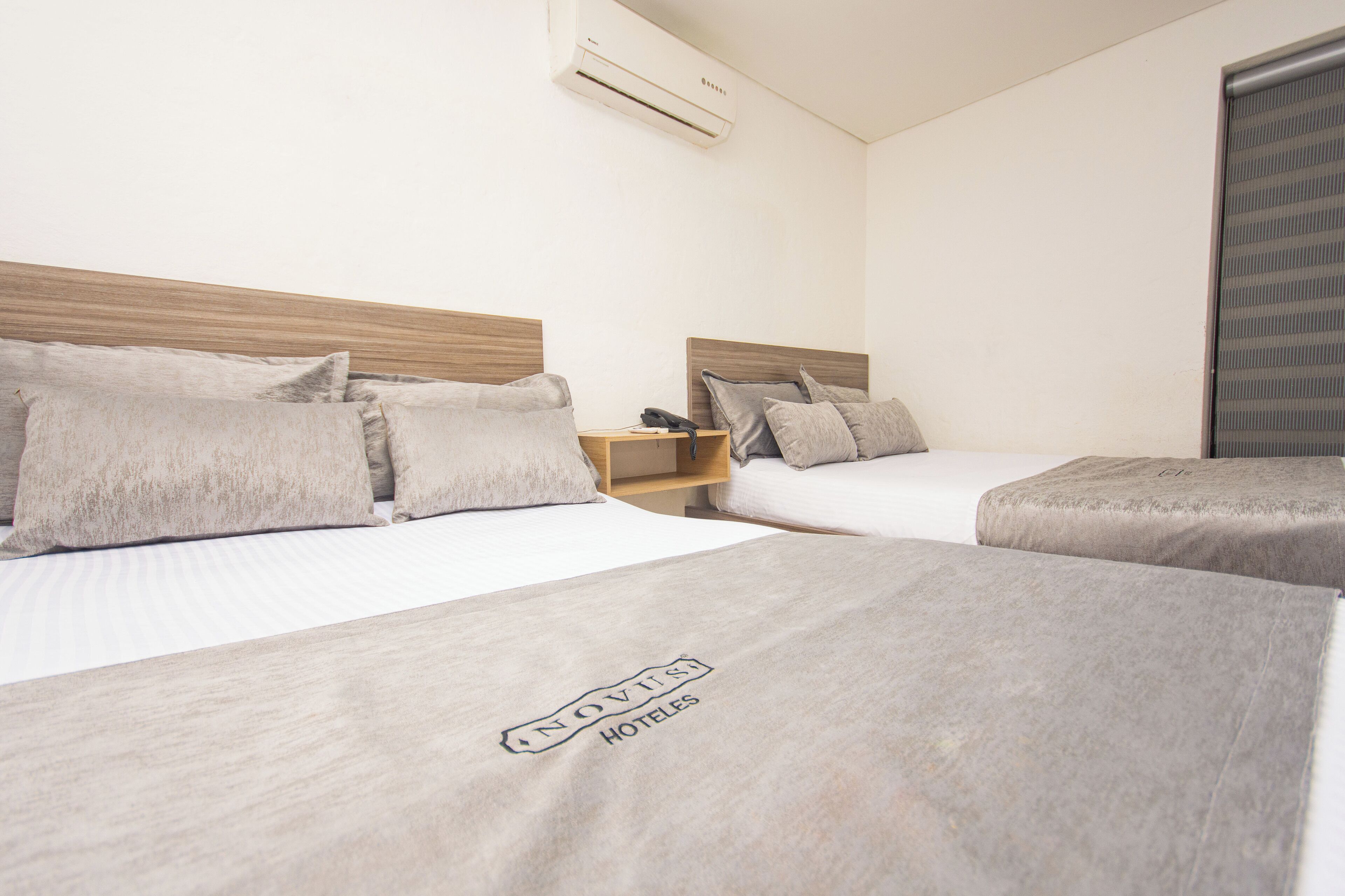 Quarto de hotel com duas camas, cada uma com cabeceira de madeira e manta cinzenta. Há uma mesa de cabeceira com abajur e telefone.