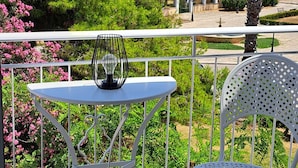 Uma varanda com mesa e duas cadeiras, com vista para um jardim com árvores e um edifício ao fundo.
