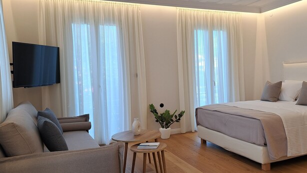 Quarto de hotel moderno com uma cama grande, televisão de tela plana na parede, área de estar com sofá e uma mesa pequena, e janelas amplas com cortinas translúcidas.