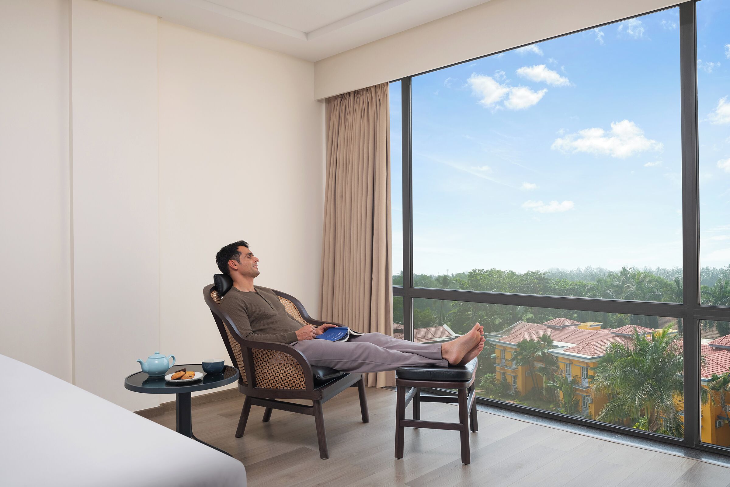 Uma pessoa relaxando em um quarto moderno com uma janela grande, uma mesinha com bule e xícara, e vista para prédios e áreas verdes lá fora.