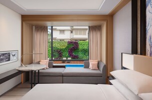 Quarto de hotel moderno com sofá, uma mesa pequena, cama e vista para um jardim através de uma janela grande.
