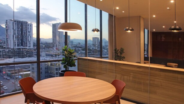 Uma zona de refeições moderna com uma mesa redonda e cadeiras laranja, grandes janelas com vista para a cidade e iluminação pendente.