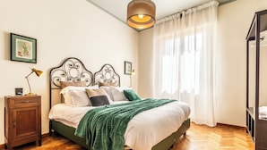 Uma cama bem arrumada com um cobertor verde, um criado-mudo de madeira com uma luminária e um quadro na parede.