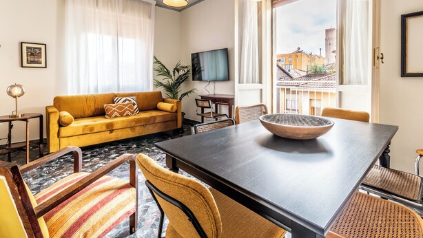 Sala de estar moderna com sofá, mesa de jantar e cadeiras. Uma televisão está montada na parede e uma grande janela oferece vista para edifícios.