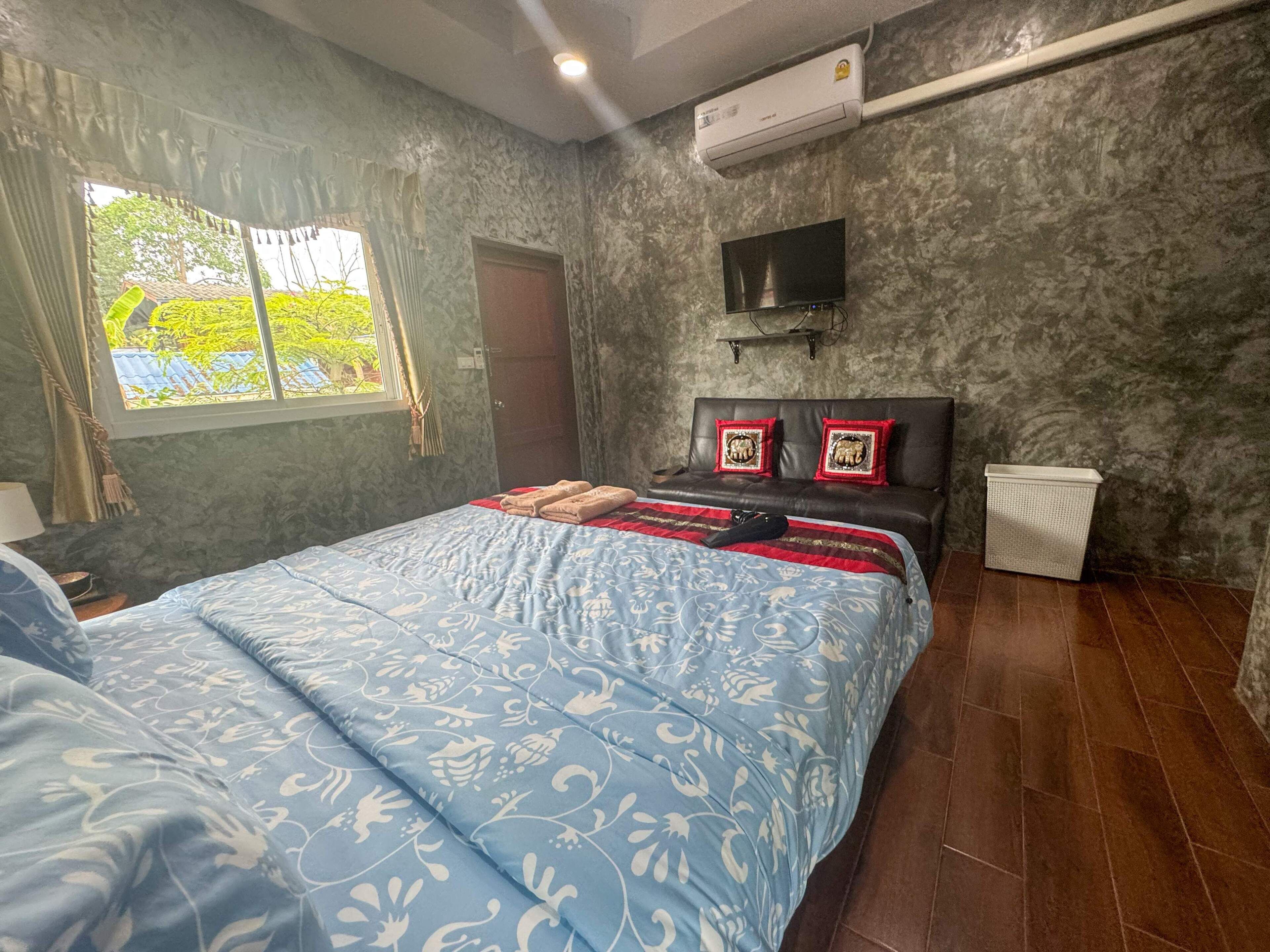 Quarto com cama, TV fixada na parede, janela com cortinas e um cesto de lixo branco.