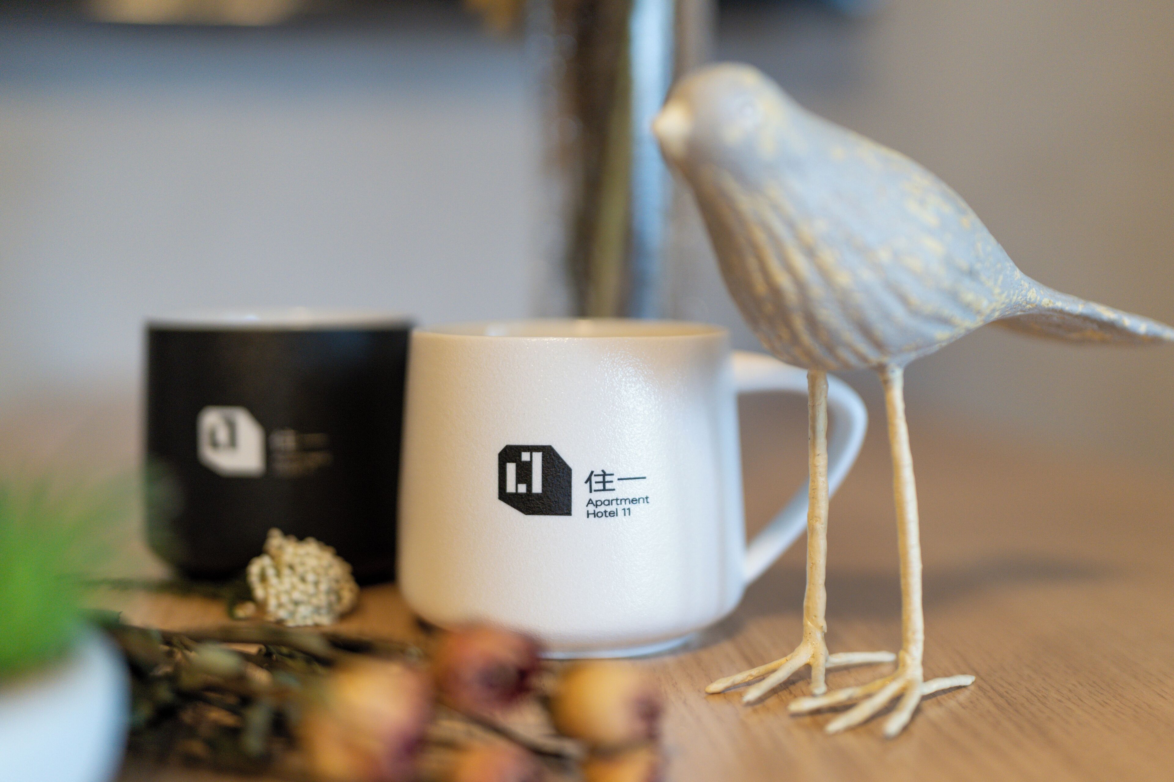 Uma caneca branca com logotipo e texto, e uma pequena estatueta de pássaro sobre uma superfície de madeira.