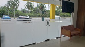 Uma área de recepção com sofá, televisão fixada na parede e vista para a rua, com carros e palmeiras.