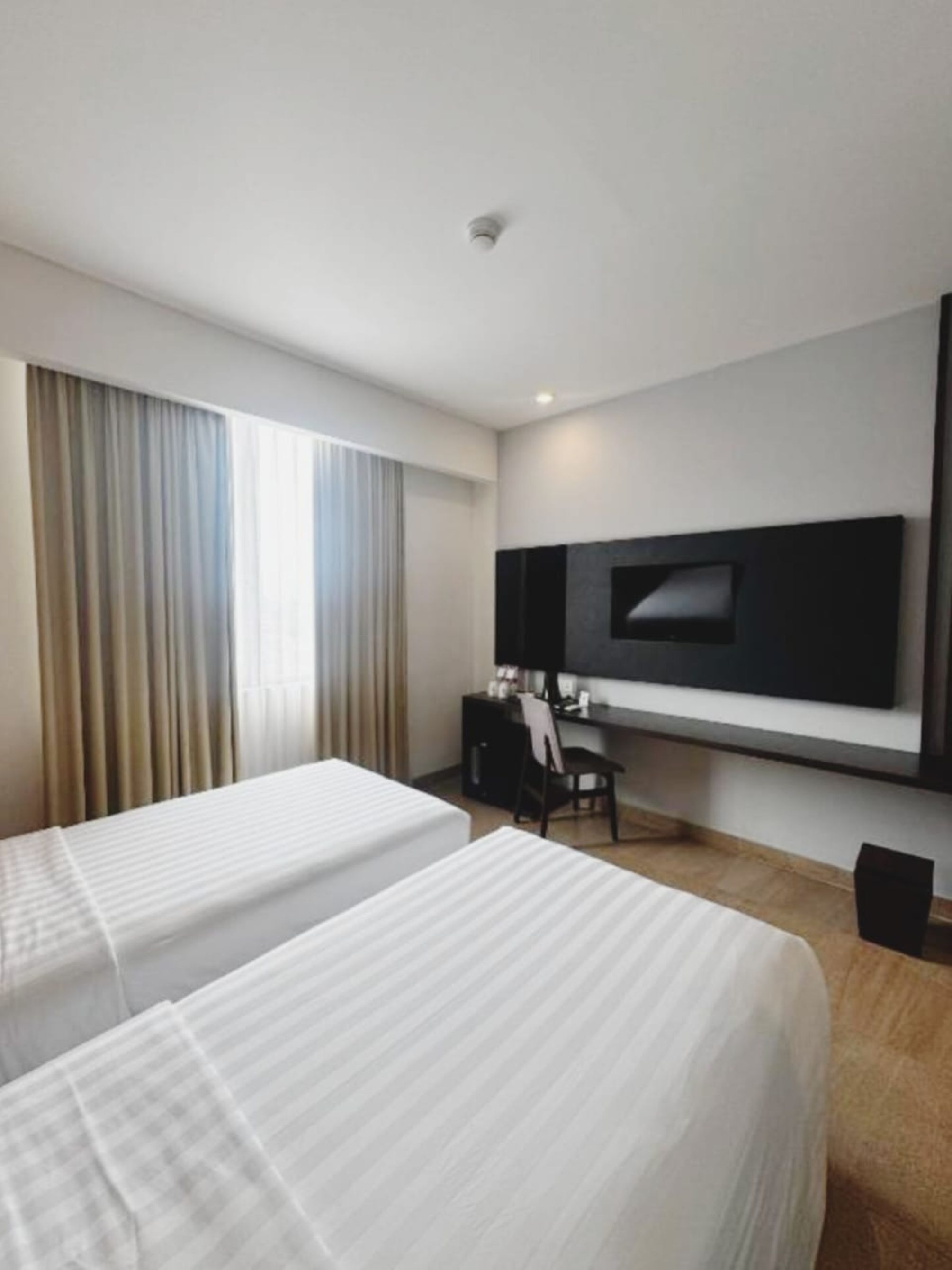Quarto de hotel com duas camas, uma escrivaninha, uma cadeira, televisão fixada na parede e janela com cortinas.