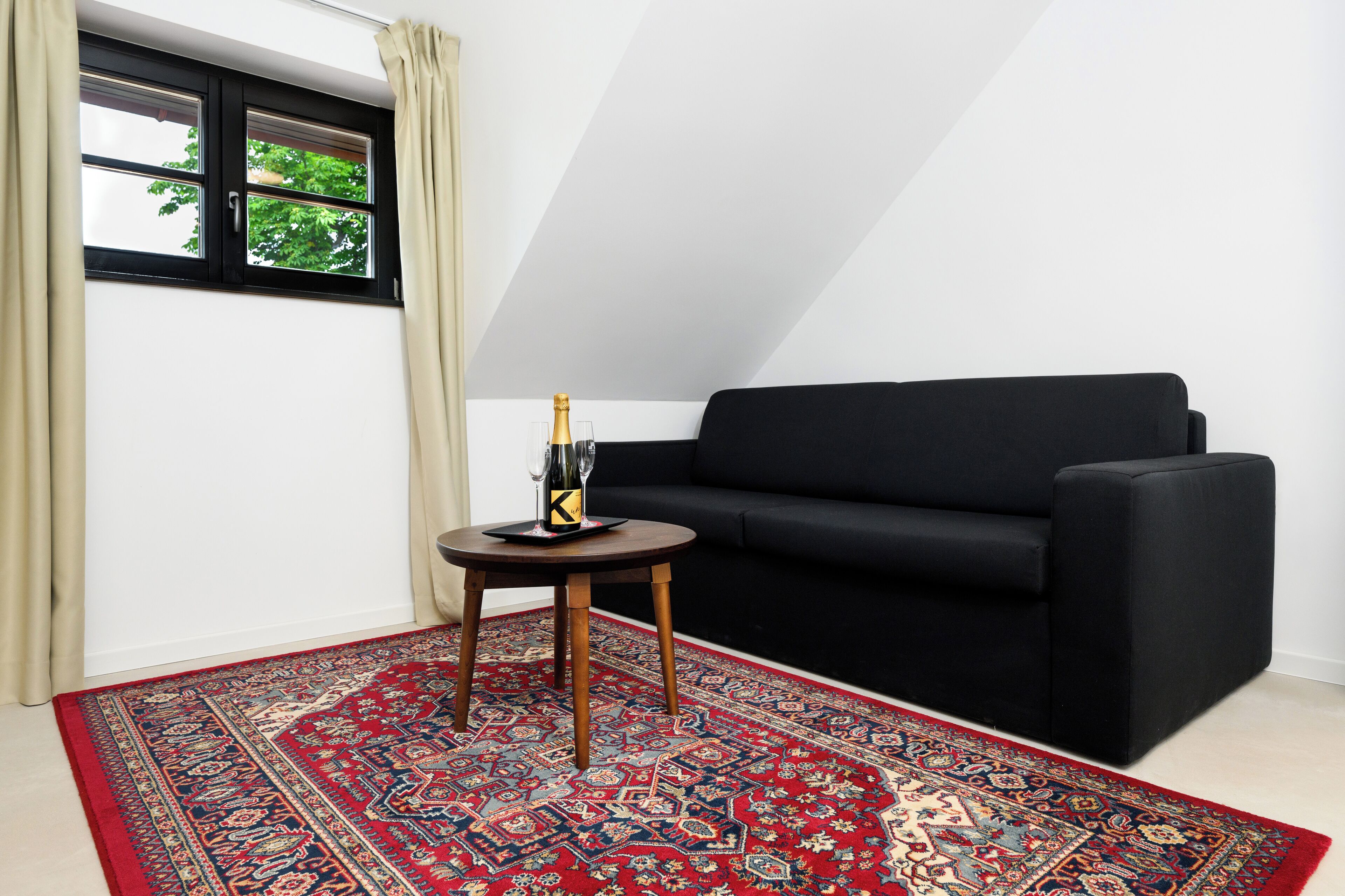 Um quarto com um sofá preto, uma pequena mesa de madeira com uma garrafa e um copo, e um tapete vermelho estampado.