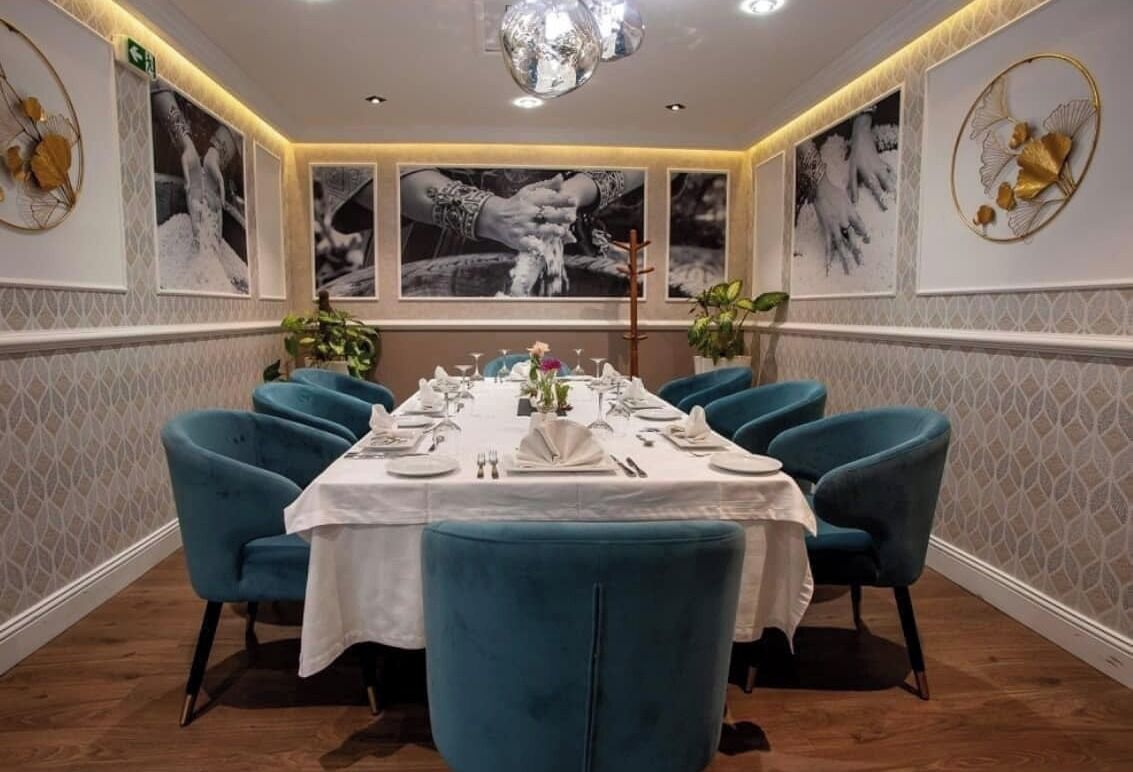 Uma sala de jantar com uma mesa comprida posta para seis pessoas, rodeada por cadeiras cor de azul-turquesa, com quadros nas paredes e um candelabro.