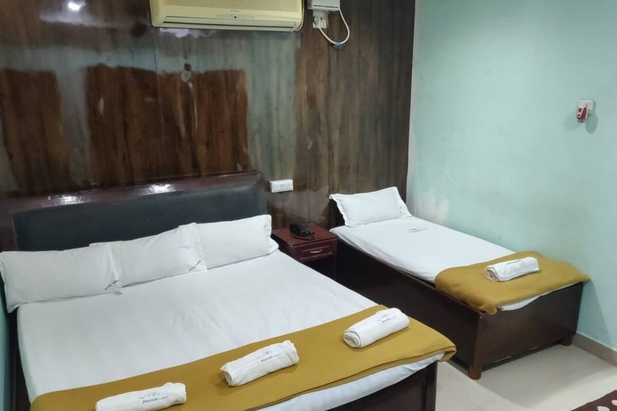 Quarto de hotel com duas camas de solteiro, cada uma com colchão branco e cabeceira marrom. Entre as camas, uma pequena mesa de cabeceira de madeira com um abajur.
