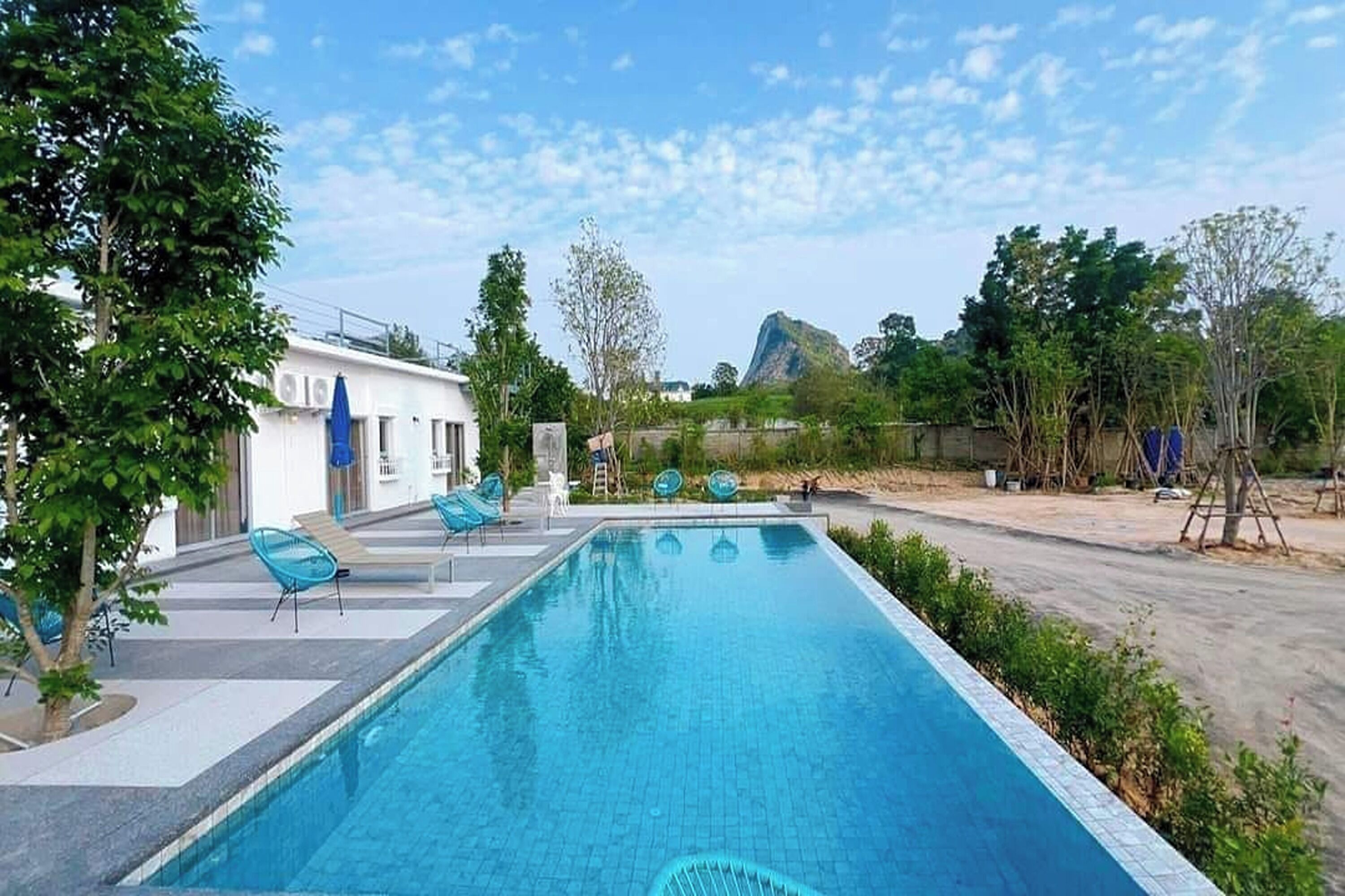 Uma piscina retangular com água azul cristalina, rodeada por um deck pavimentado. Há várias espreguiçadeiras e um pequeno edifício com fachada branca ao fundo.