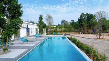 Uma piscina retangular com água azul cristalina, rodeada por um deck pavimentado. Há várias espreguiçadeiras e um pequeno edifício com fachada branca ao fundo.