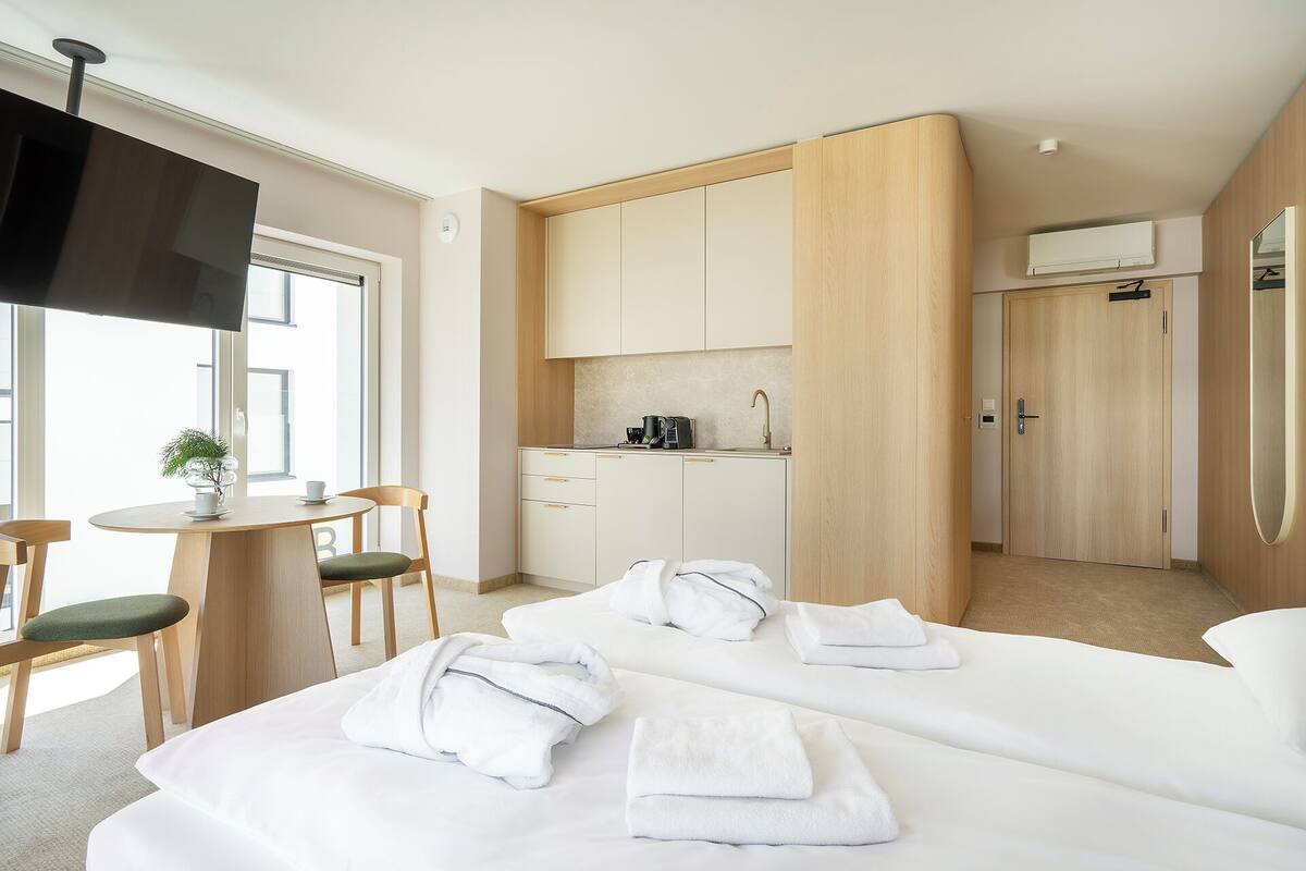 Quarto de hotel com cama, escrivaninha, cadeira, mesa pequena, planta e cozinha compacta.