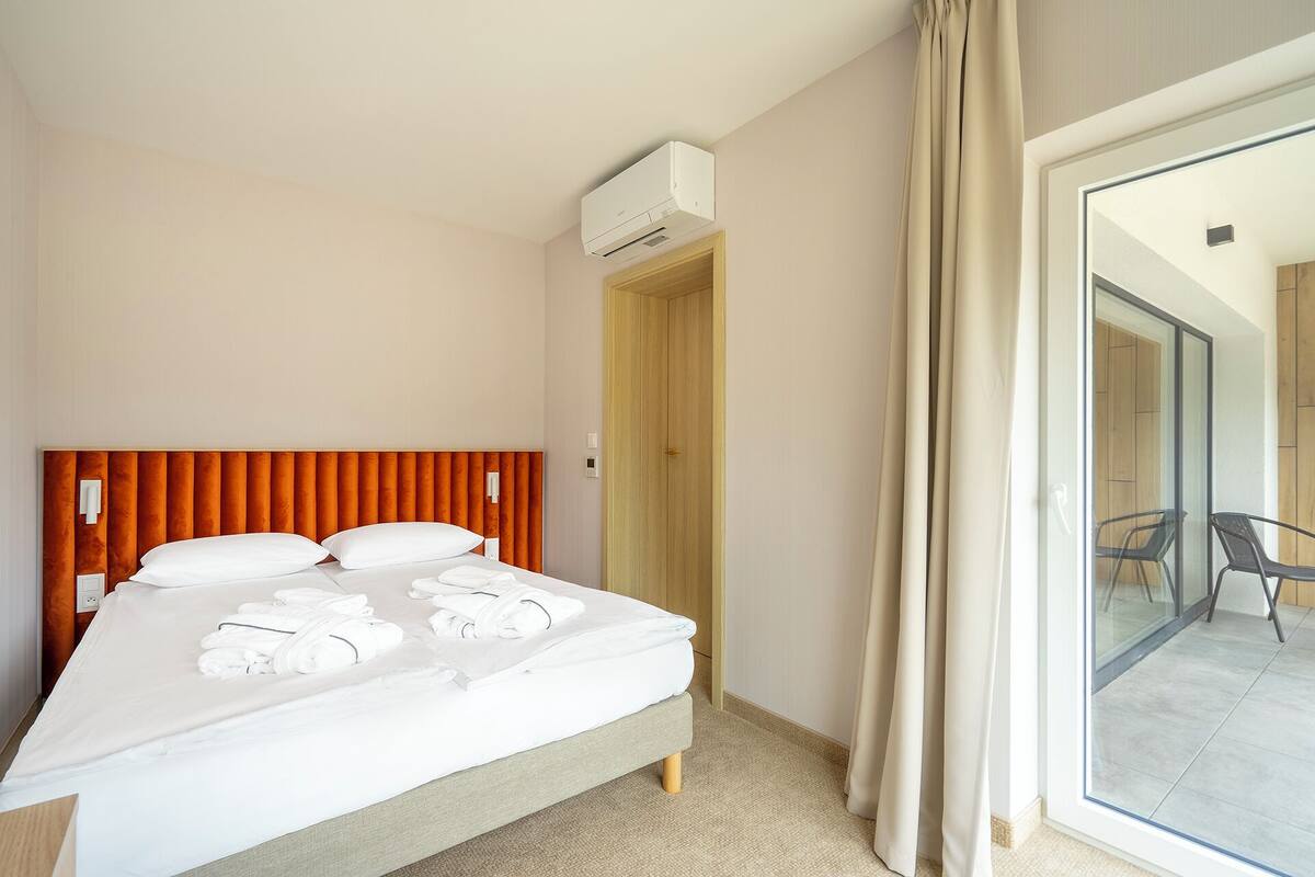 Quarto de hotel com cama, cabeceira de madeira, ar condicionado e porta de vidro deslizante para uma varanda, além de uma cadeira lá fora.