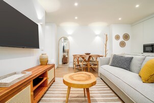 Uma sala de estar moderna com uma mesa de centro de madeira, um sofá com almofadas, uma TV na parede, uma área de jantar com mesa e cadeiras, e uma cozinha com micro-ondas.