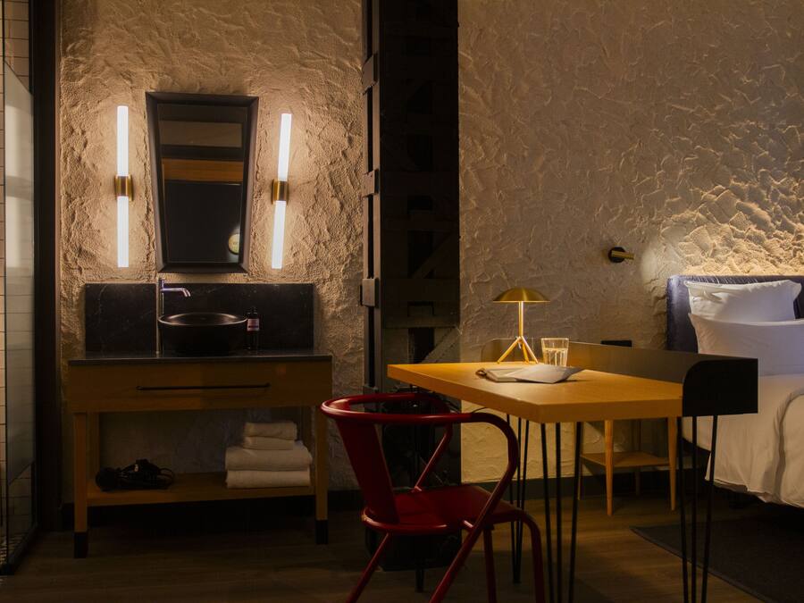 Quarto de hotel compacto com uma cama, uma escrivaninha, uma cadeira, uma mesinha, um espelho e uma prateleira com uma toalha e um sapato.