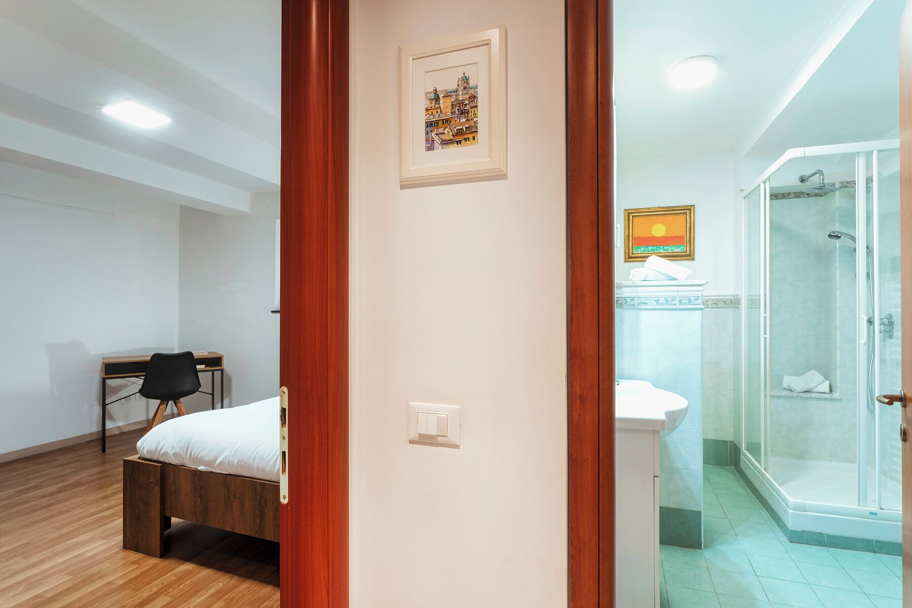 Quarto de hotel com cama de madeira, uma escrivaninha, uma cadeira, banheiro com chuveiro e um quadro na parede.