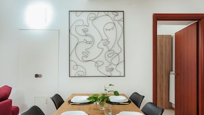 Uma sala de jantar com uma mesa de madeira posta para quatro pessoas, cercada por cadeiras pretas, uma poltrona vermelha e uma obra de arte abstrata emoldurada na parede.