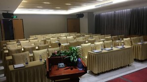 Uma sala de conferências com fileiras de cadeiras bege, uma mesa comprida com um microfone e um tapete vermelho.