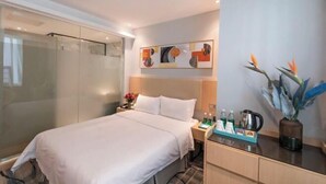 Quarto de hotel com cama, mesa de cabeceira, obra de arte na parede, um vaso com flores e box de chuveiro de vidro.