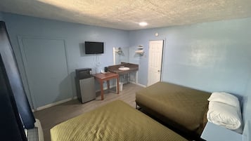 Quarto de hotel pequeno e individual, com duas camas de solteiro, uma escrivaninha com cadeira, televisão na parede e um refrigerador.