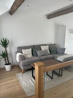 Uma sala de estar moderna com um sofá seccional cinza, uma mesa de centro de madeira, um vaso de planta e uma grande janela com persianas.