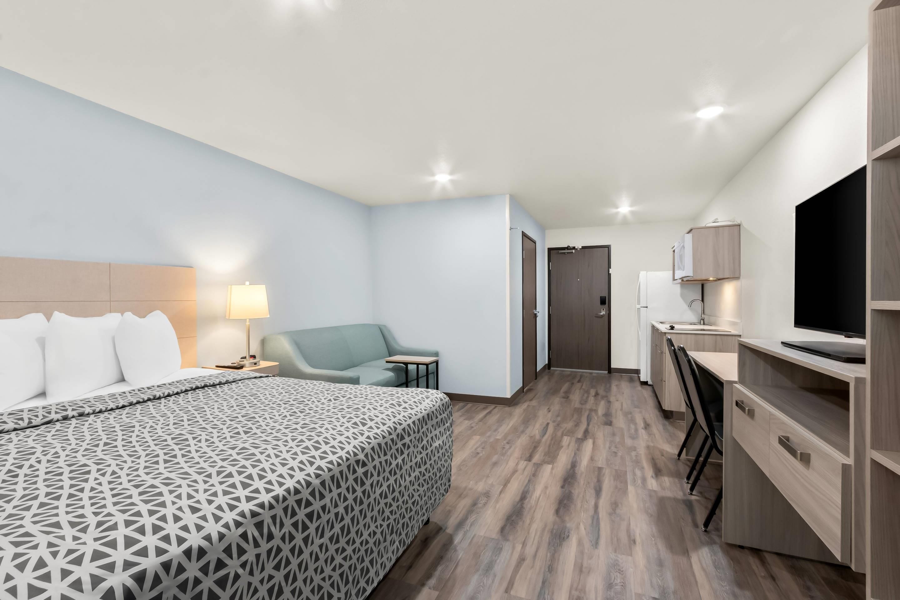 Quarto de hotel com cama, escrivaninha, cadeira, televisão e cozinha compacta.