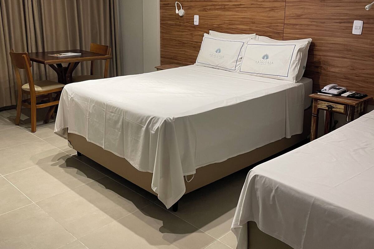 Quarto de hotel com uma cama grande, uma cama menor, uma mesa de cabeceira de madeira, uma mesa com cadeiras e uma cortina.