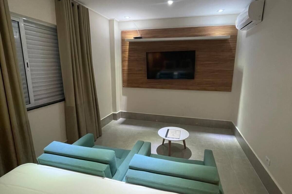 Quarto de hotel moderno com uma cama grande, uma mesa redonda pequena e uma TV fixada num painel de madeira.
