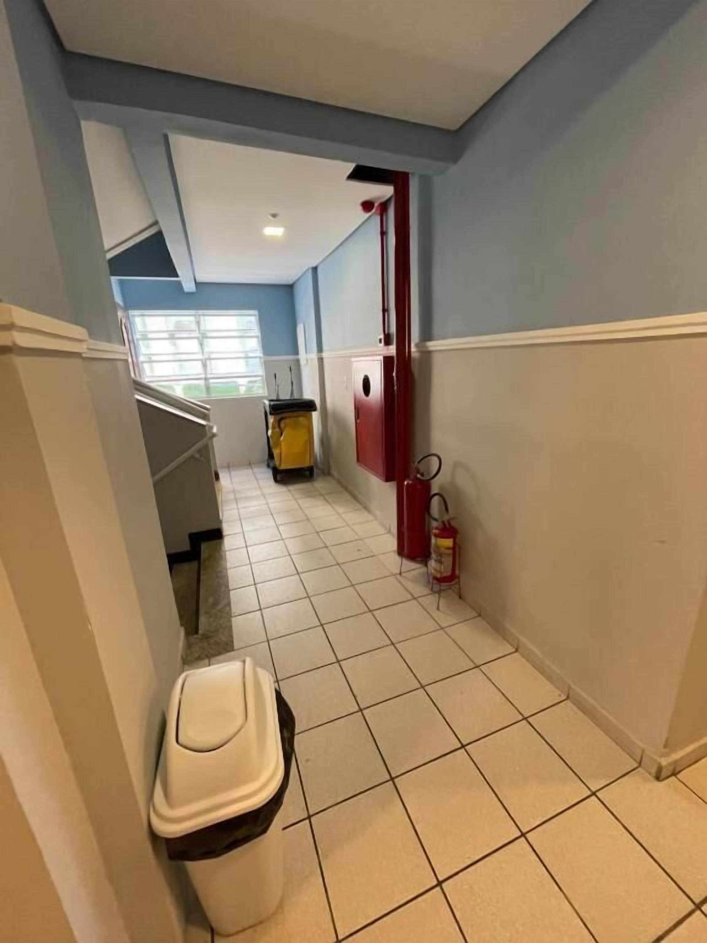 Um corredor estreito com piso de azulejos, um extintor de incêndio vermelho e um carrinho de limpeza amarelo.