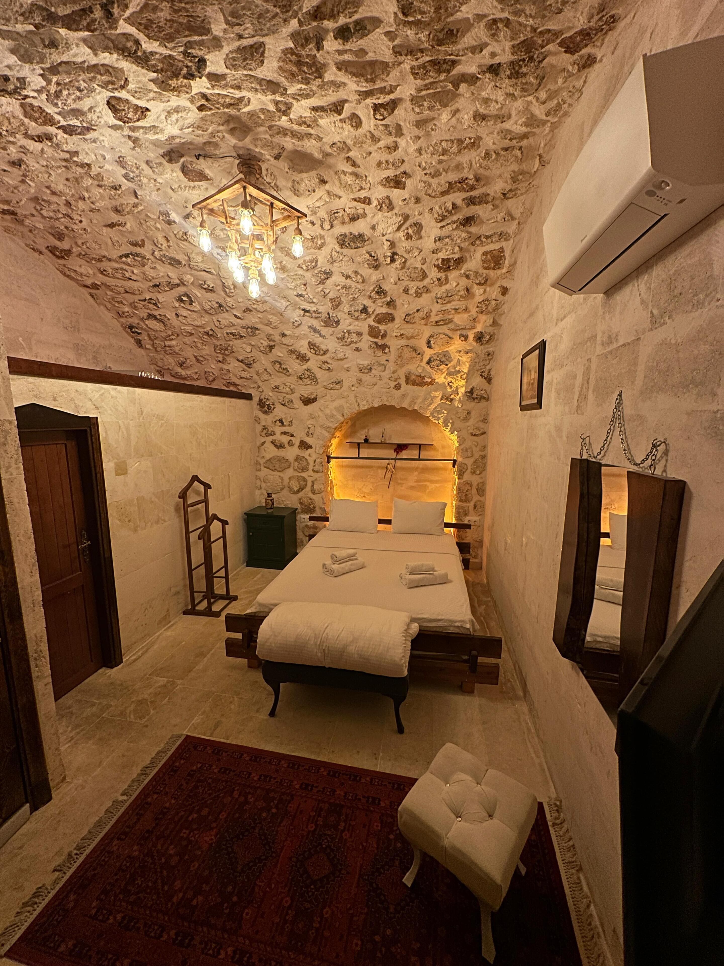 Quarto com paredes de pedra, uma cama, um banco, um espelho e um lustre.