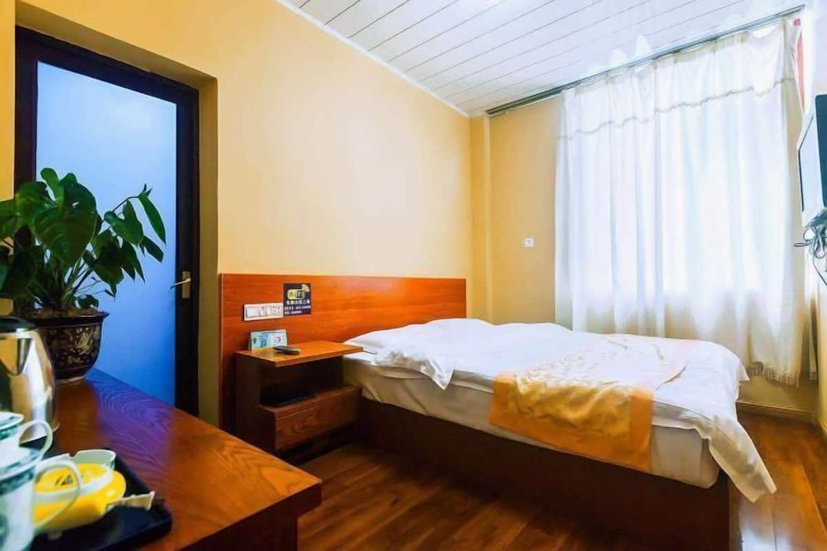 Quarto de hotel com cama, mesa de cabeceira de madeira, planta em vaso, bandeja com itens de café da manhã e janela com cortinas.