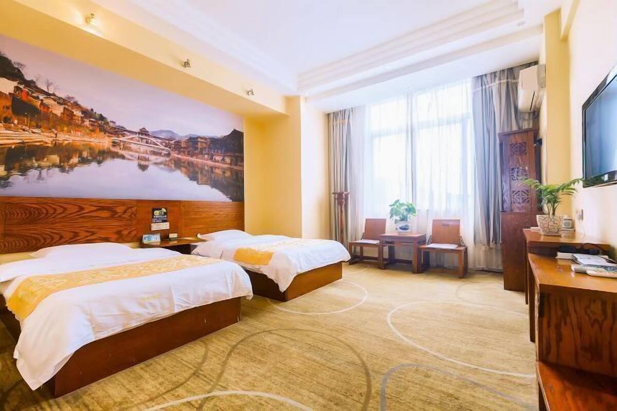Quarto de hotel com duas camas, uma escrivaninha, uma cadeira, televisão e um quadro grande na parede.