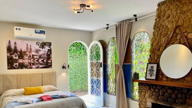 Quarto com cama, parede de pedra, lareira e uma janela grande com vista para o verde.