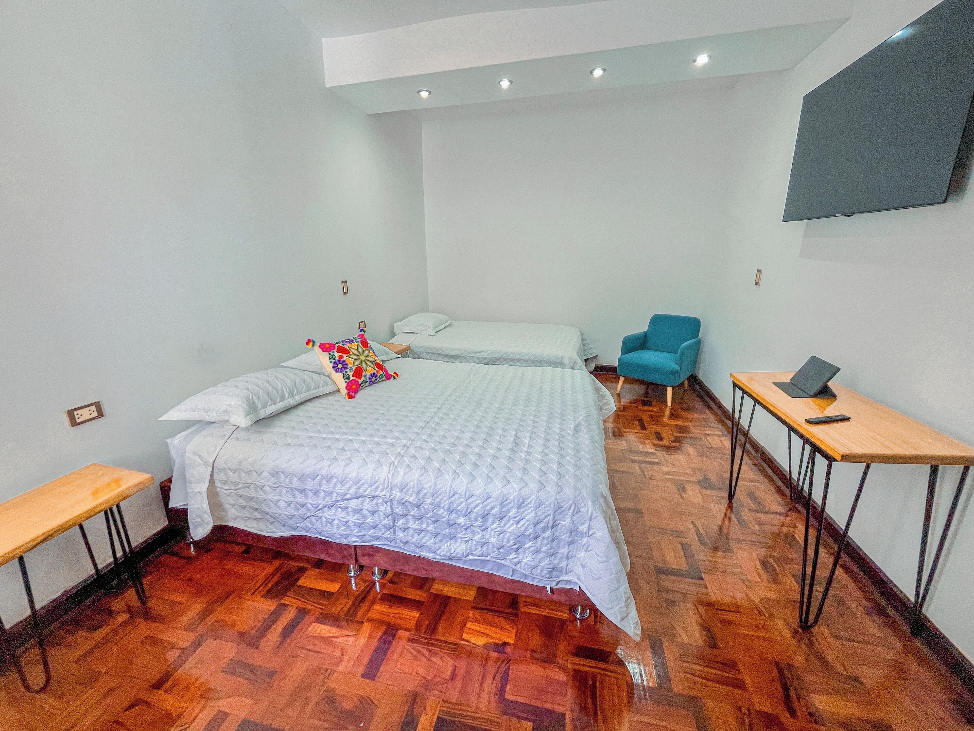Quarto com duas camas, uma cadeira azul, uma mesinha e uma televisão de tela plana na parede.
