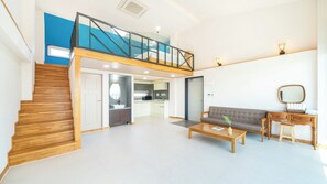 Ampla sala de estar com escada, sofá, mesa de centro e cozinha ao fundo.