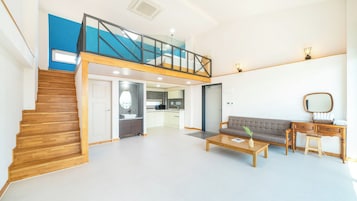 Ampla sala de estar com escada, sofá, mesa de centro e cozinha ao fundo.