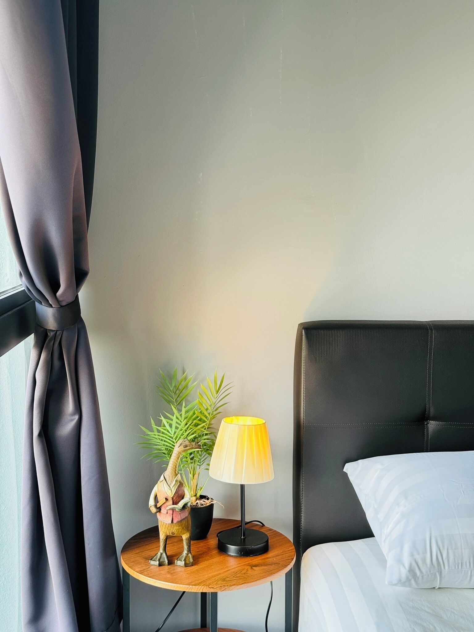 Cama arrumada com travesseiro branco e manta listrada, uma pequena mesa redonda de madeira com luminária e planta, e cortina escura parcialmente aberta.