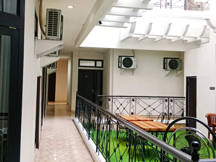 Um corredor com piso de azulejos, uma mesa de madeira com cadeiras, uma área com grama sintética verde e um corrimão de metal.