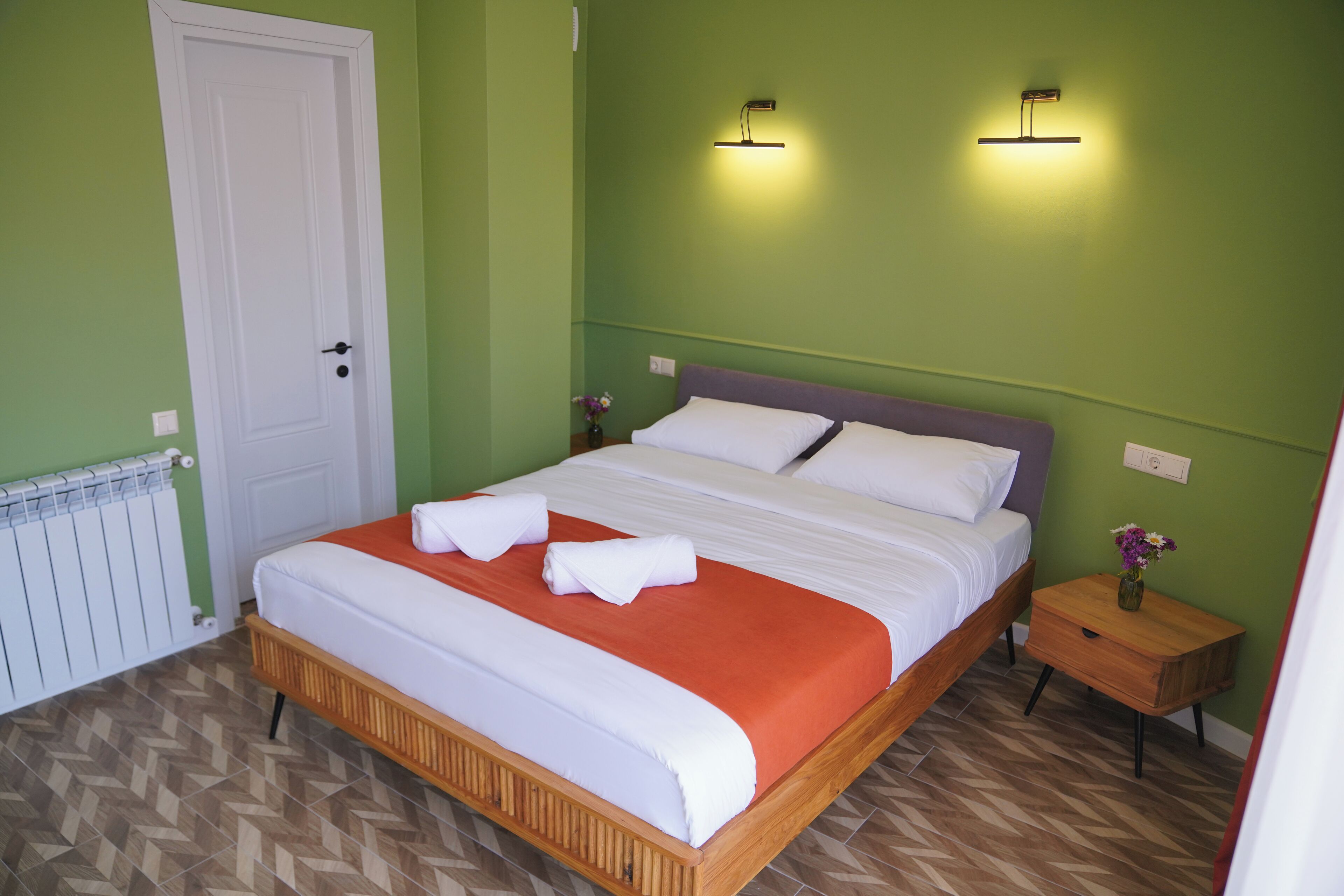 Uma cama bem arrumada com lençóis brancos e um cobertor laranja, uma mesinha de cabeceira de madeira com um vaso de flores, e um radiador na parede.
