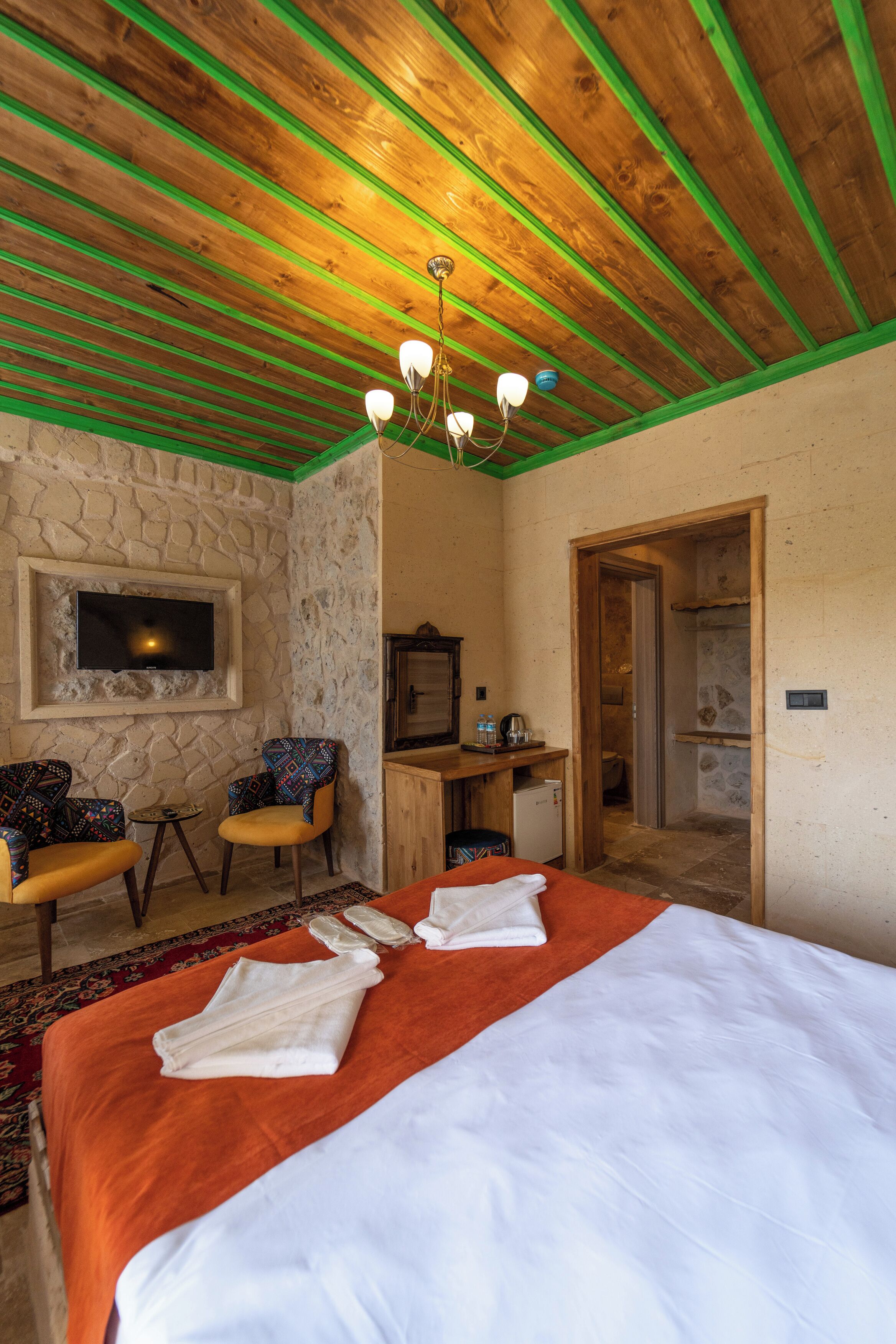 Quarto com cama, duas cadeiras, escrivaninha, televisão e parede de pedra.